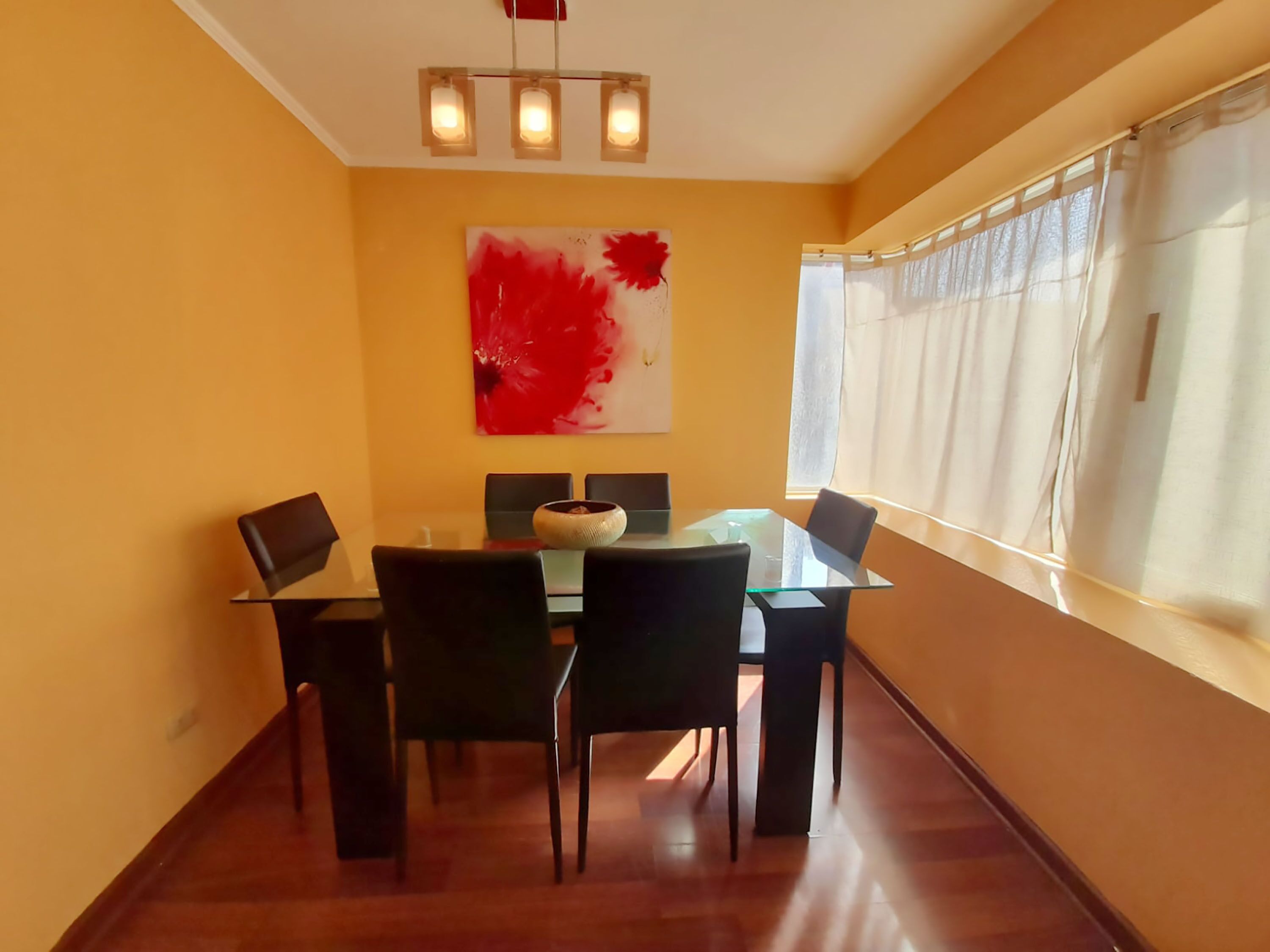 Uma sala de jantar com mesa de vidro, cadeiras pretas, pintura abstrata vermelha e janela com cortinas leves.
