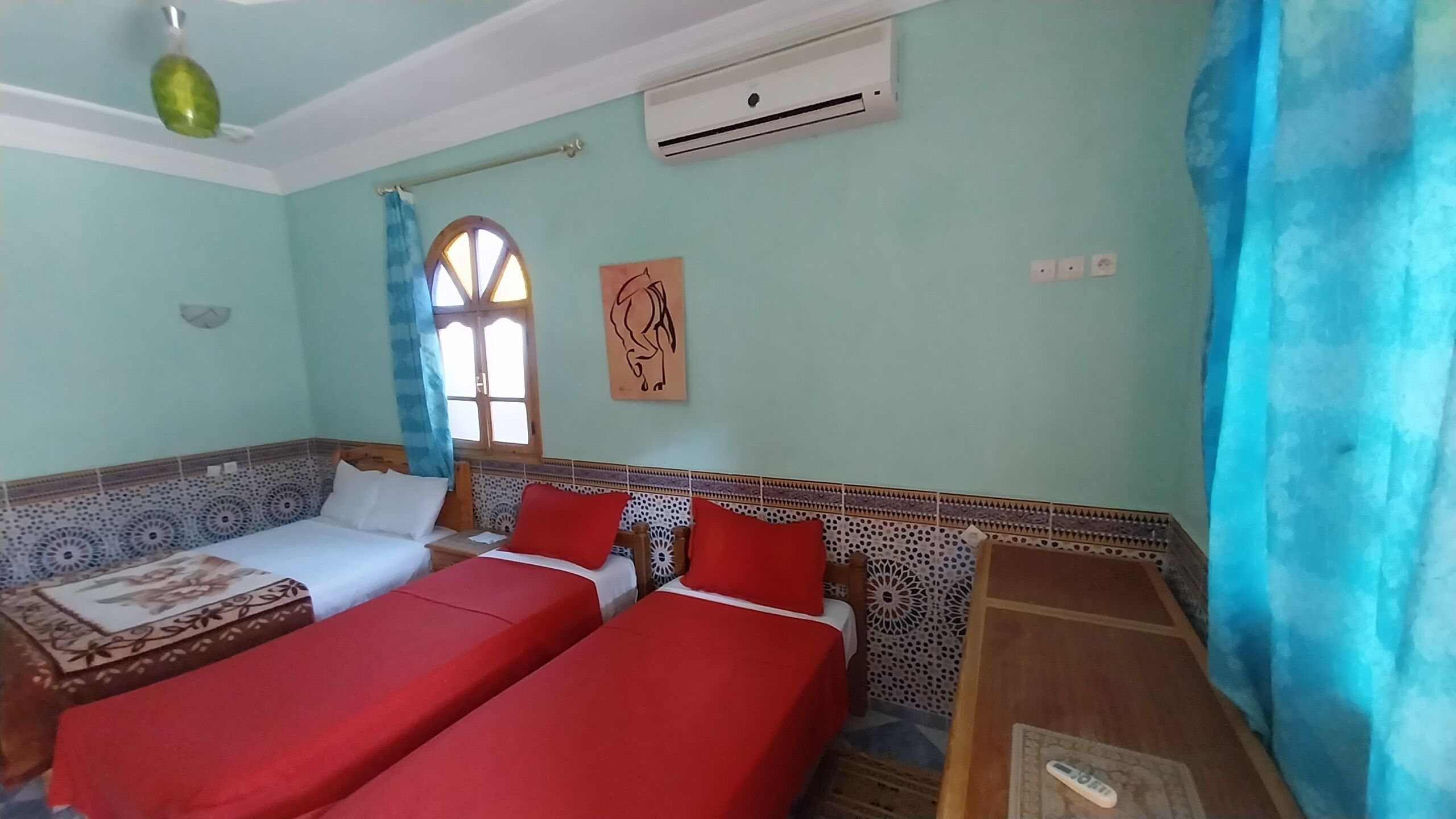 Quarto com duas camas, janela com cortinas azuis, ar condicionado fixado na parede e um objeto decorativo na parede.
