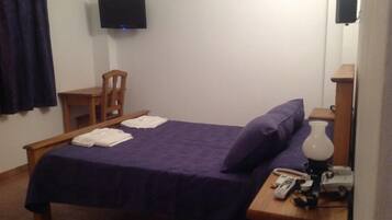 Quarto pequeno e simples com uma cama, uma cadeira, uma escrivaninha e uma televisão na parede.