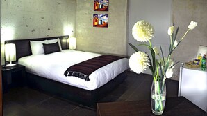Quarto de hotel com cama, mesas de cabeceira, um abajur, um vaso com flores e duas pinturas nas paredes.