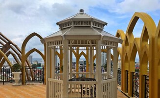 Um gazebo branco com telhado pontiagudo, rodeado por arcos amarelos, com vista para uma paisagem urbana com edifícios e um corpo de água.