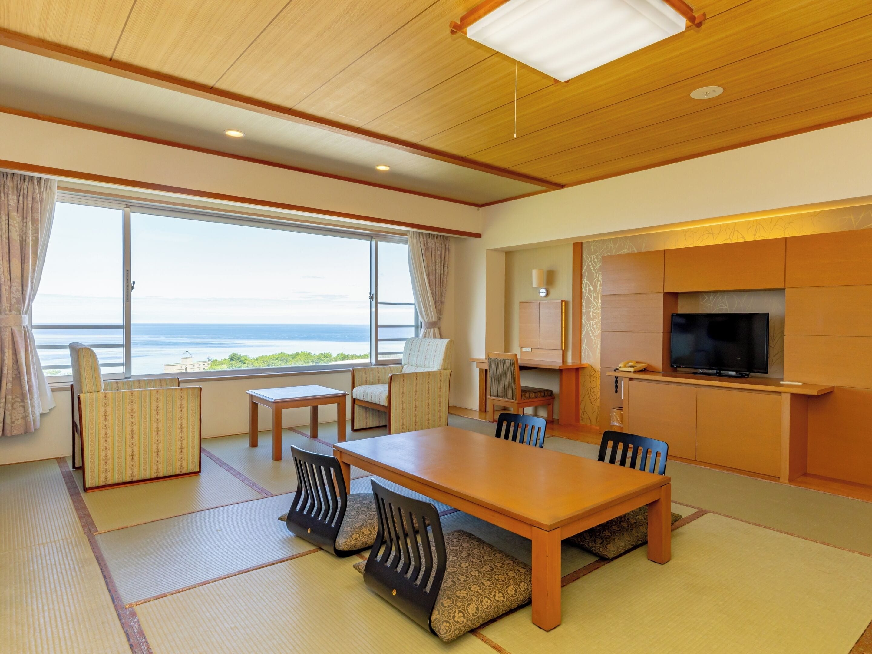 Um quarto espaçoso com uma grande mesa de jantar de madeira, cadeiras estofadas, televisão e vista para o mar.