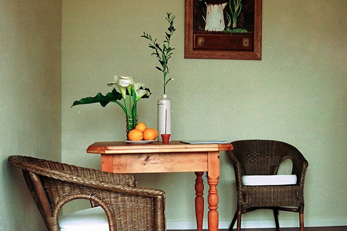 Uma área de refeições com mesa de madeira, duas cadeiras de vime, um vaso com flores e um quadro na parede.