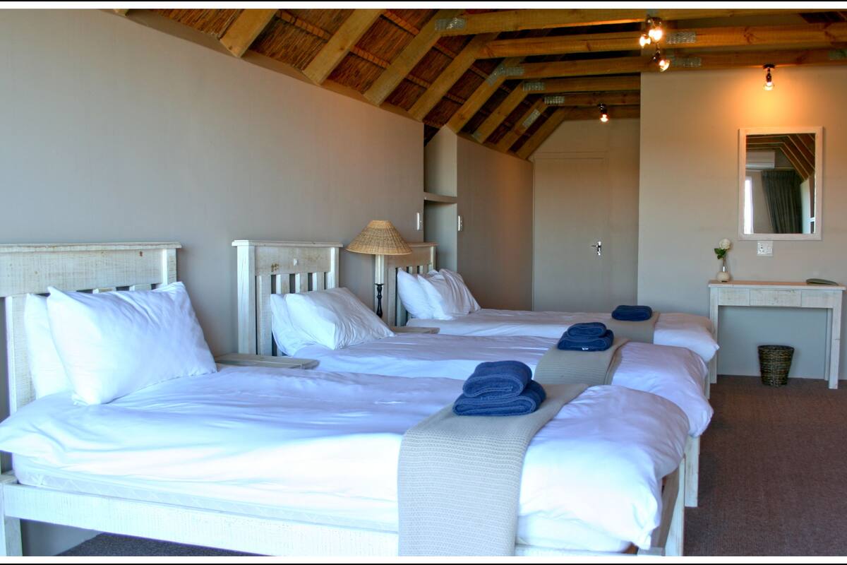 Quarto de hotel com duas camas de solteiro, uma pequena escrivaninha com espelho e teto com vigas de madeira aparentes.