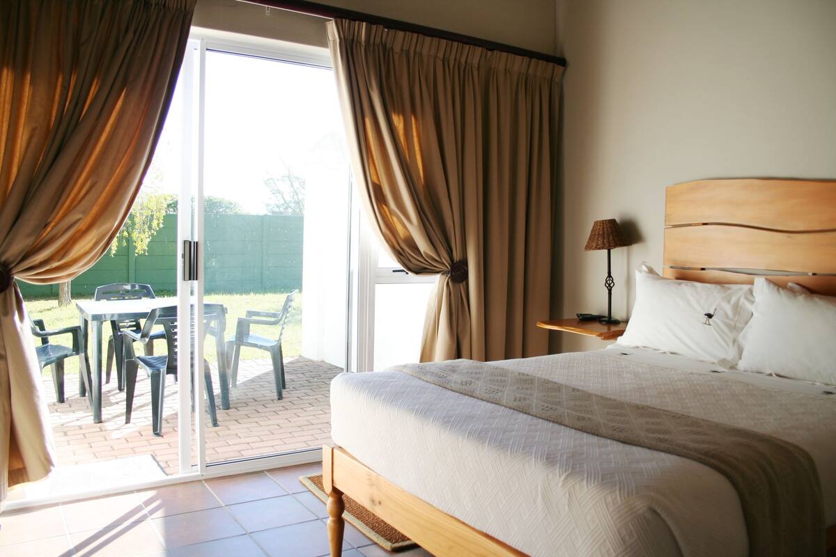 Quarto com uma cama grande, cabeceira de madeira, mesa de cabeceira com abajur e vista para um pátio externo com cadeiras e mesa.