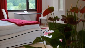 Quarto com cama, escrivaninha, cadeira e janela com vista para uma floresta.