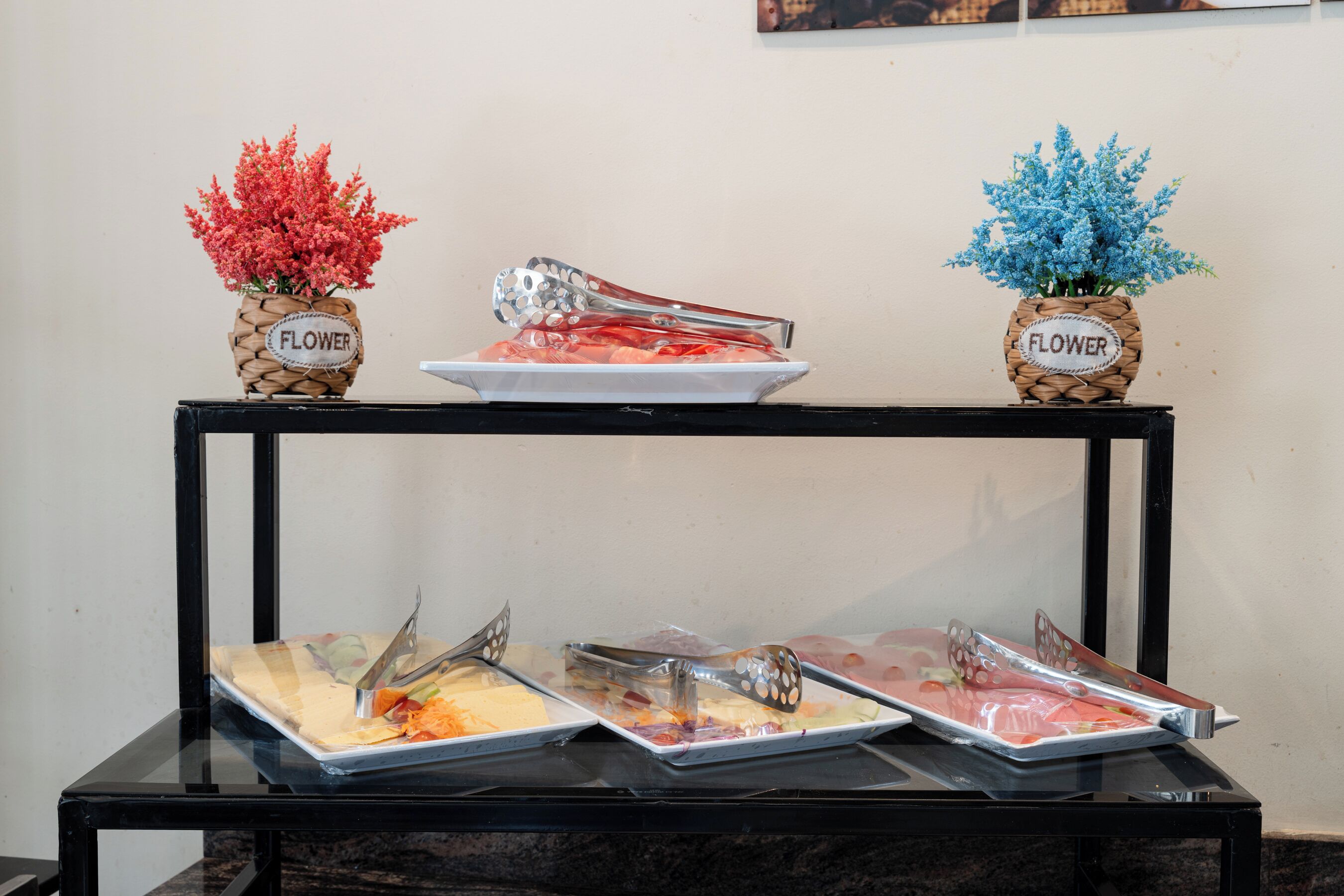 Montagem de buffet com dois vasos rotulados "FLOWER" contendo flores artificiais, e três bandejas de comida com utensílios de servir.