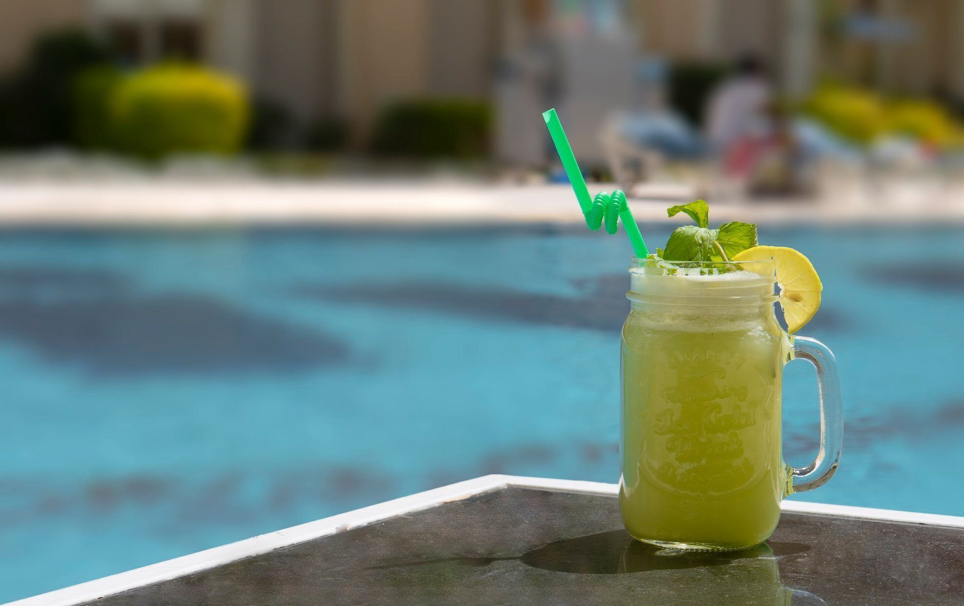 Um copo de vidro com canudo verde e folhas de hortelã, apoiado sobre uma mesa perto de uma piscina.