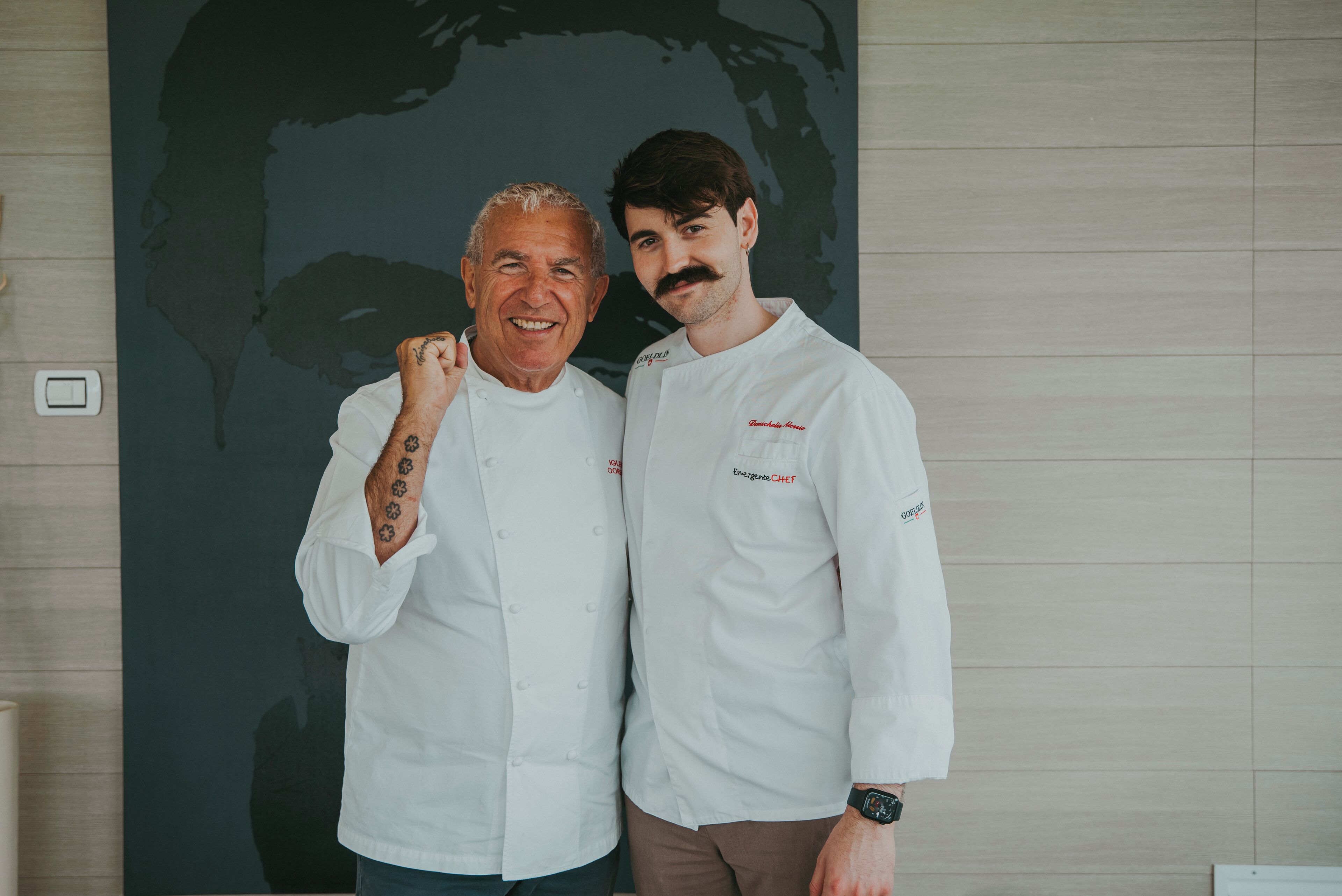 Duas pessoas vestidas com uniformes de chef em frente a um grande mural de um mapa-múndi.
