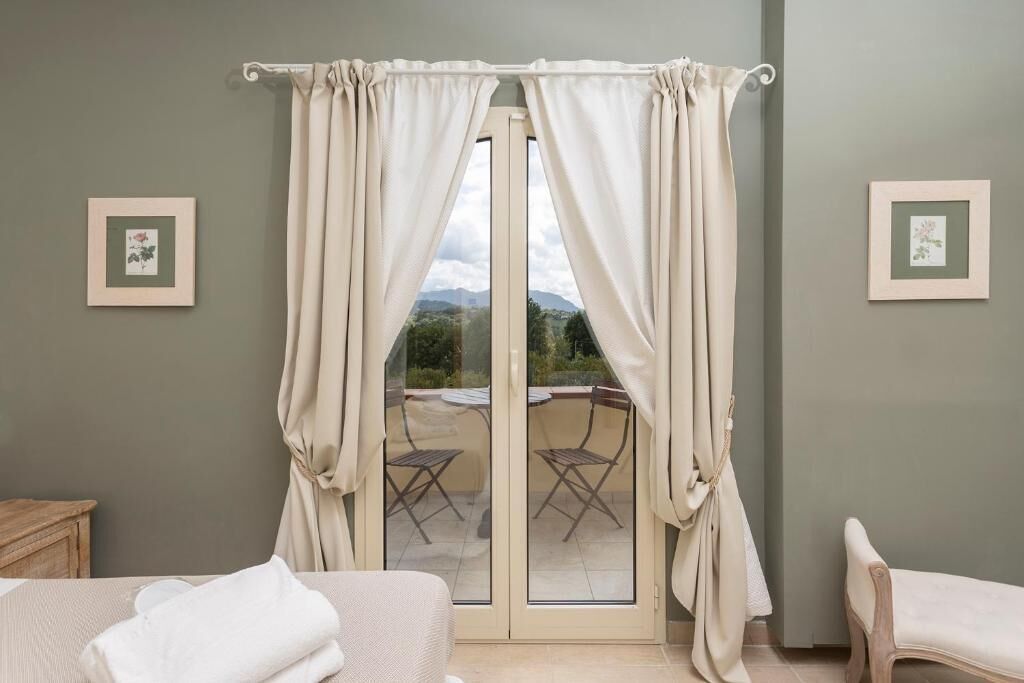 Quarto com porta de vidro deslizante que dá para um pátio externo, cortinas bege, uma cadeira de madeira e quadros nas paredes.