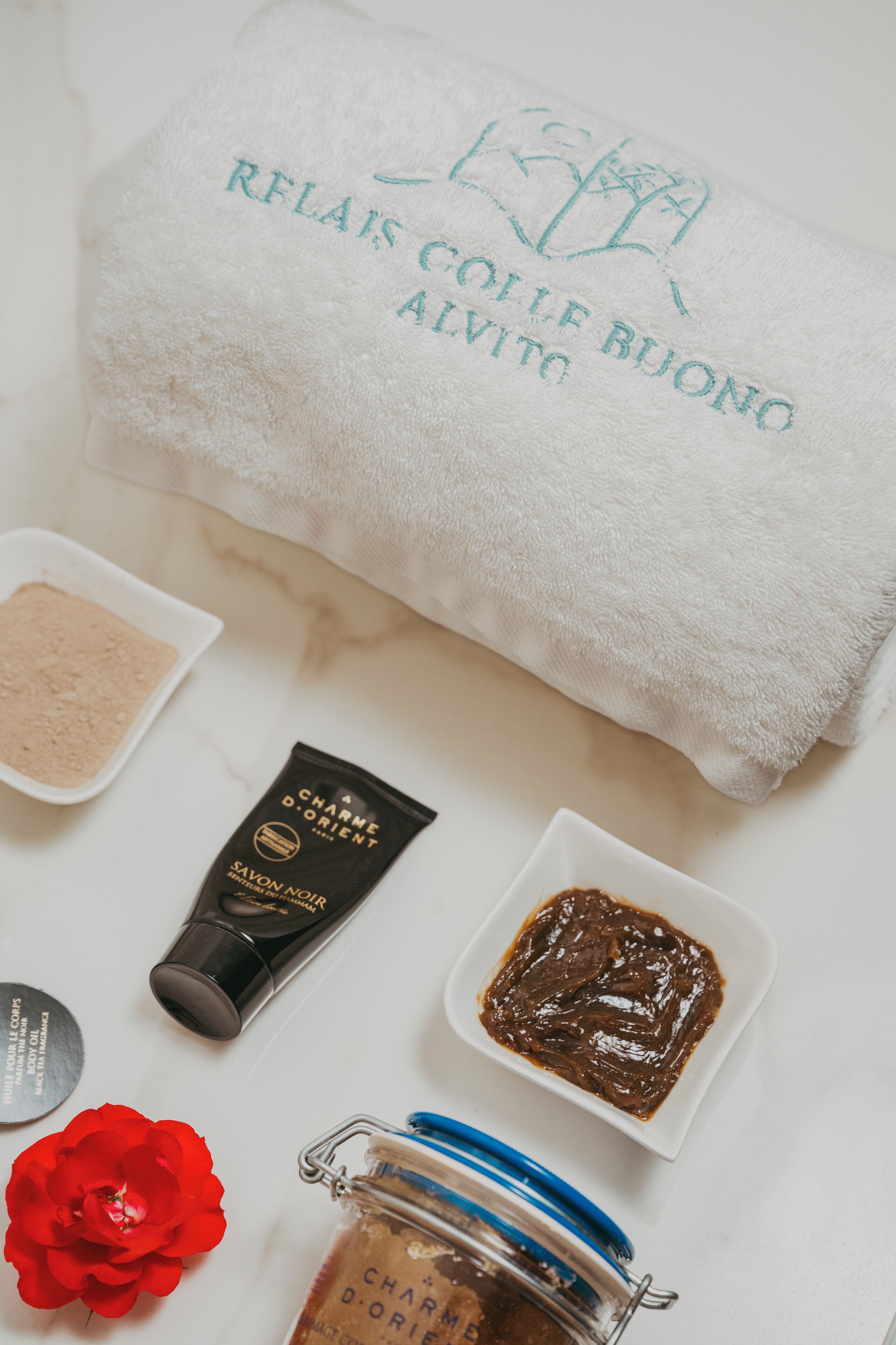 Toalha branca dobrada com texto bordado, um pequeno frasco de creme, um tubo de creme, uma pequena tigela de pó e uma flor vermelha sobre uma superfície branca.