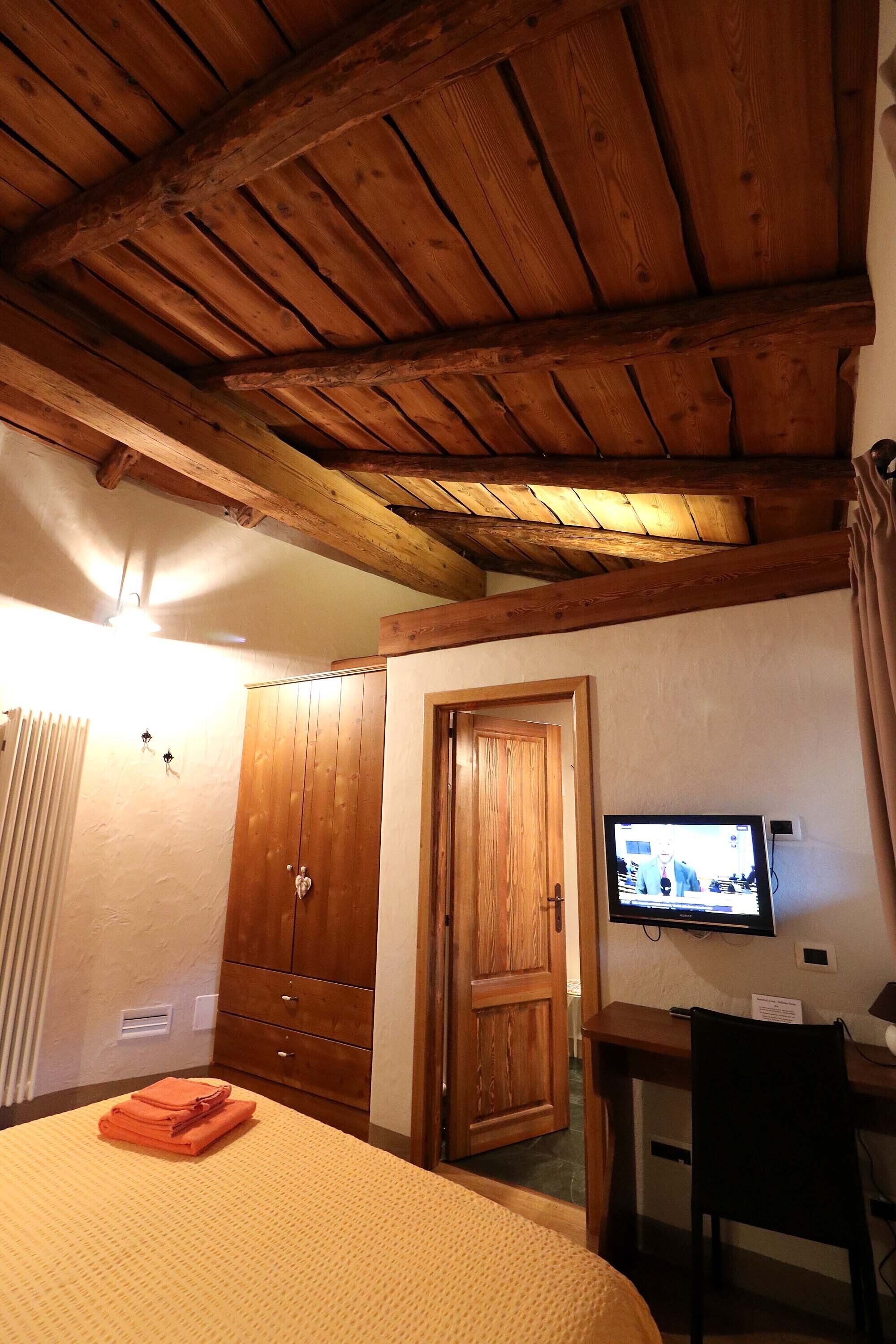 Quarto com teto de madeira, cama com colcha amarela, guarda-roupa de madeira, televisão sobre uma mesa e porta que dá para outro cômodo.