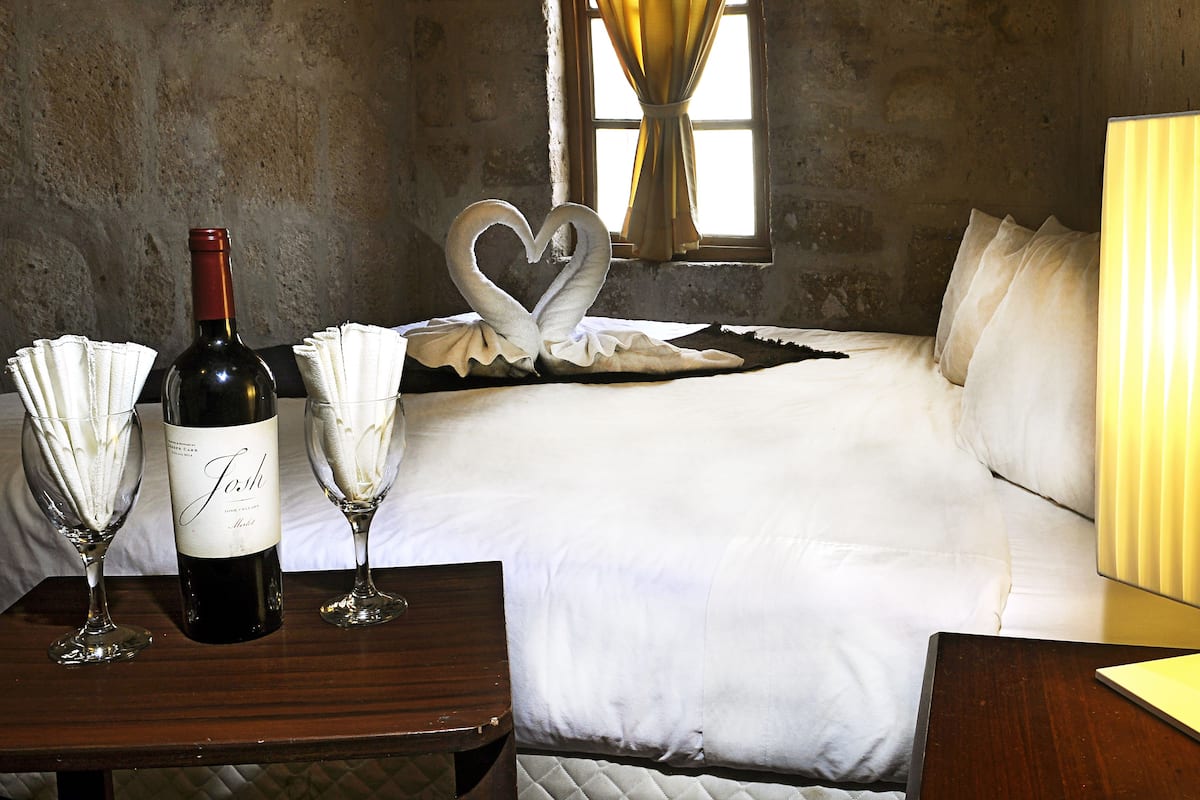 Quarto de hotel com uma cama, uma garrafa de vinho e duas taças de vinho sobre uma mesa de madeira.