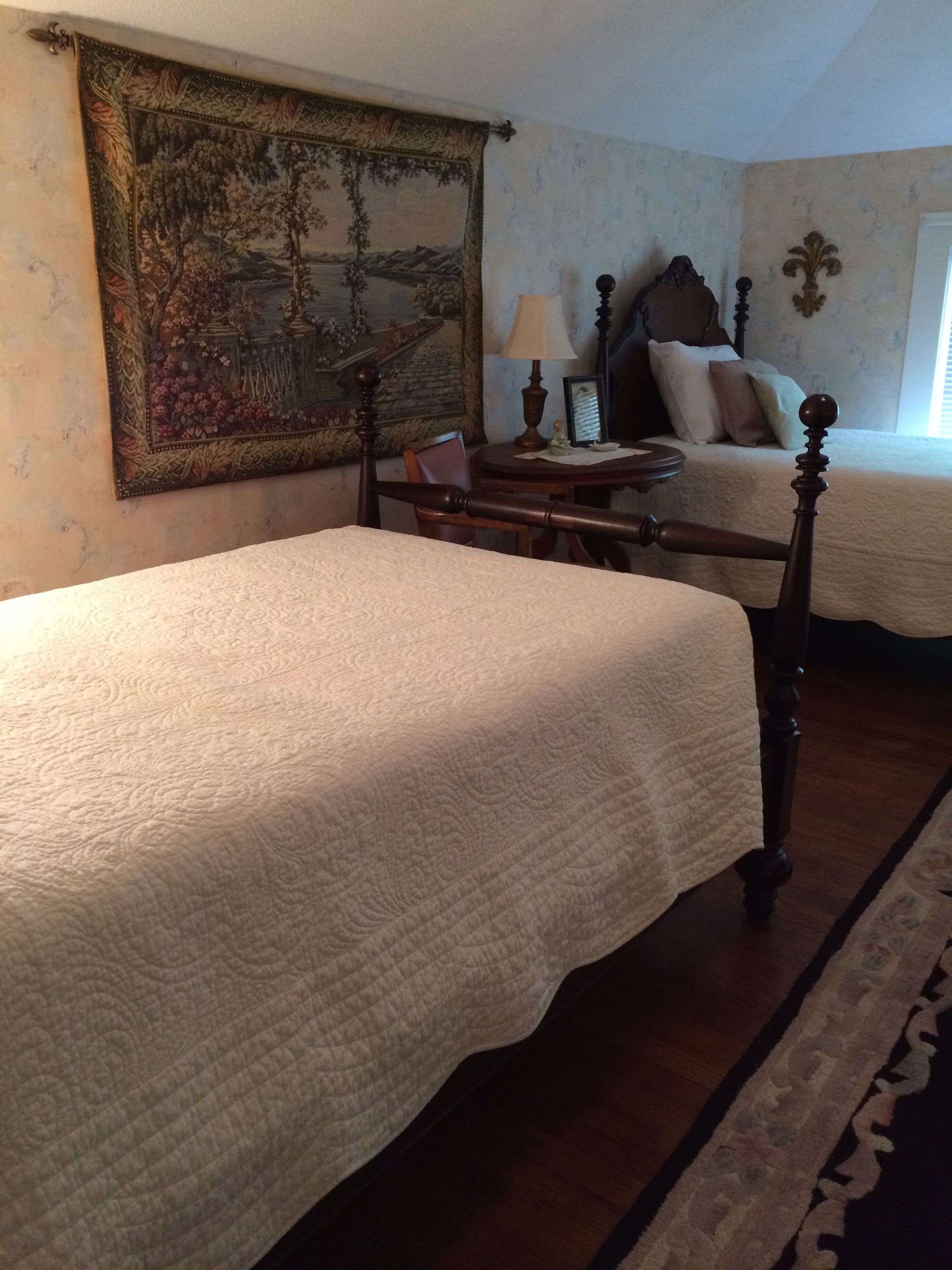 Quarto com uma cama grande, uma cama menor, uma mesa de cabeceira, um abajur e uma tapeçaria na parede.
