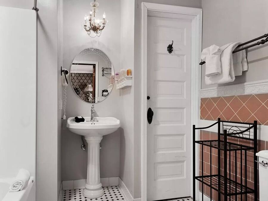 Um banheiro com pia em pedestal, espelho redondo, lustre, porta branca e porta-toalhas preto.