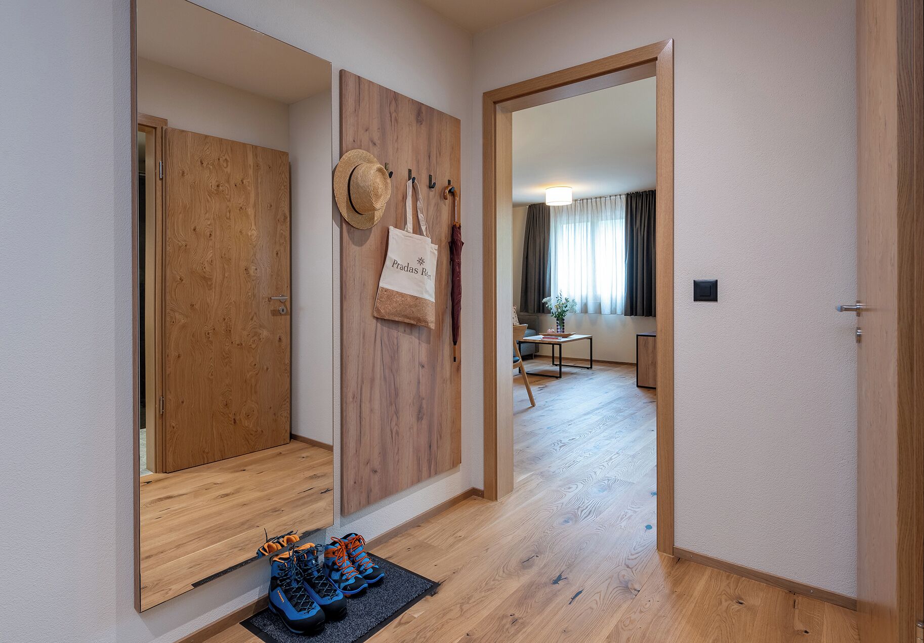 Corredor com piso de madeira, espelho, porta e vista para uma sala de estar com sofá e mesa.