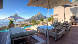Terraço com espreguiçadeiras e guarda-sóis com vista para uma piscina e montanhas.