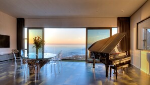 Quarto espaçoso com um piano de cauda, uma mesa de vidro e cadeiras, com vista para uma paisagem costeira deslumbrante.
