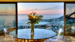 Mesa de vidro redonda com um vaso de flores e duas cadeiras transparentes, com vista para uma paisagem urbana e oceano ao pôr do sol.