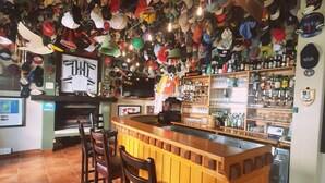 Um bar com balcão de madeira e banquetas, rodeado por prateleiras repletas de garrafas e copos.