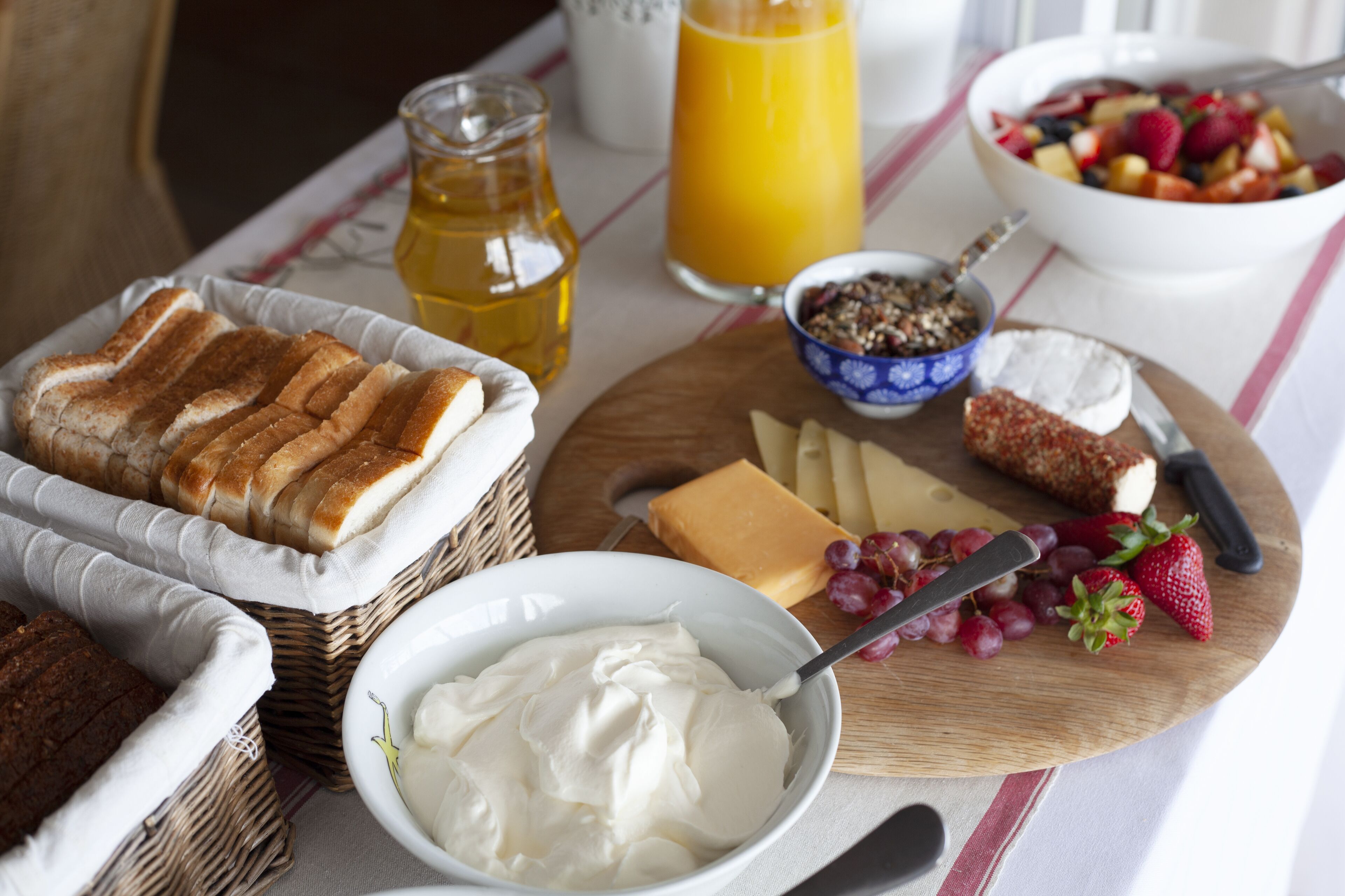 Mesa de café da manhã com pão, queijo, uvas, morangos, iogurte e salada de frutas.