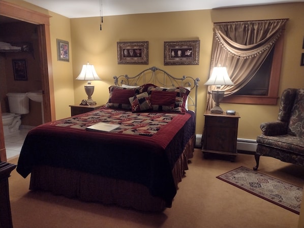 Quarto com uma cama, duas mesas de cabeceira, uma cadeira, uma janela com cortinas e uma porta que dá para um banheiro.