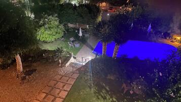 Vista noturna de um jardim com paisagismo, piscina iluminada em azul, caminho de pedra e mobiliário de exterior.