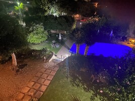 Vista noturna de um jardim com paisagismo, piscina iluminada em azul, caminho de pedra e mobiliário de exterior.