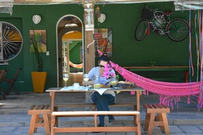 Uma pessoa sentada em uma mesa de madeira ao ar livre, com uma rede, uma bicicleta e uma parede verde com decorações.