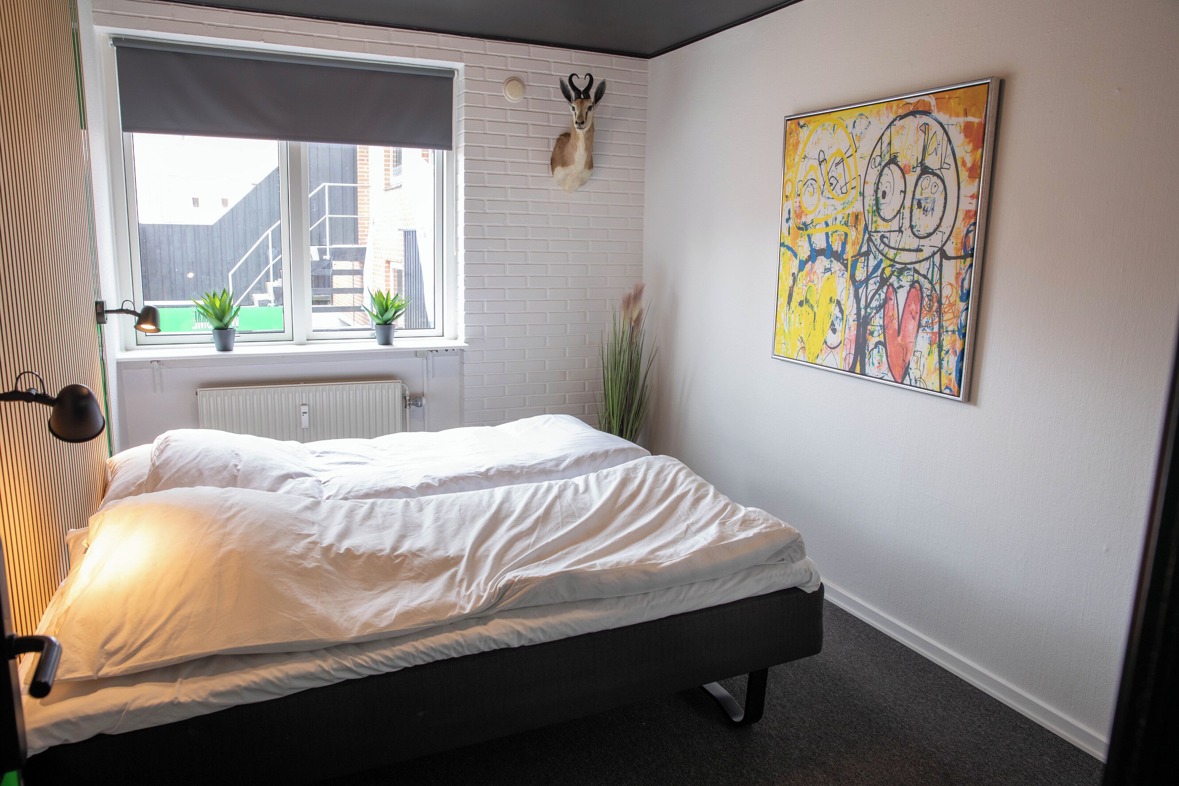 Quarto com cama, janela, radiador, cabeça de veado fixada na parede e uma pintura abstrata colorida.