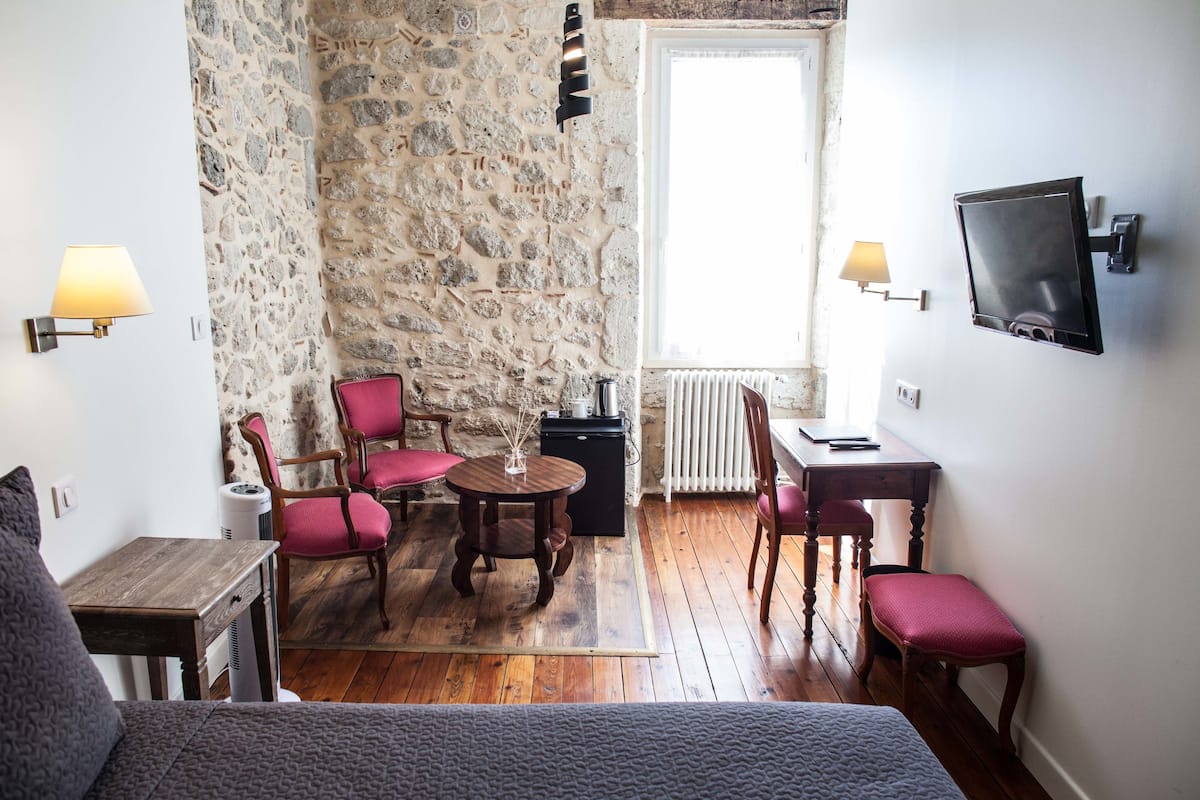 Um quarto aconchegante com parede de pedra, uma mesa redonda de madeira, duas cadeiras, uma mesinha com abajur e uma TV fixada na parede.