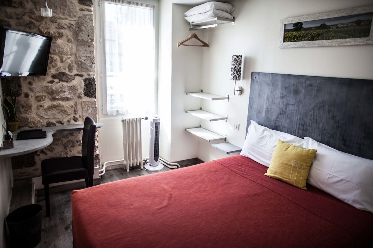 Quarto com parede de pedra, cama com roupa de cama vermelha e branca, televisão, radiador e janela com cortinas.