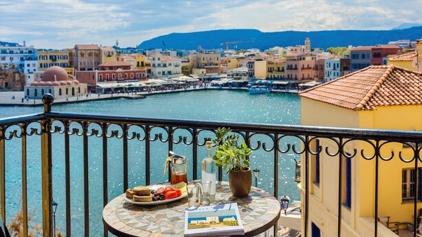 Varanda com mesa posta, comida, um livro e um vaso de planta, com vista para uma orla com barcos e edifícios coloridos.