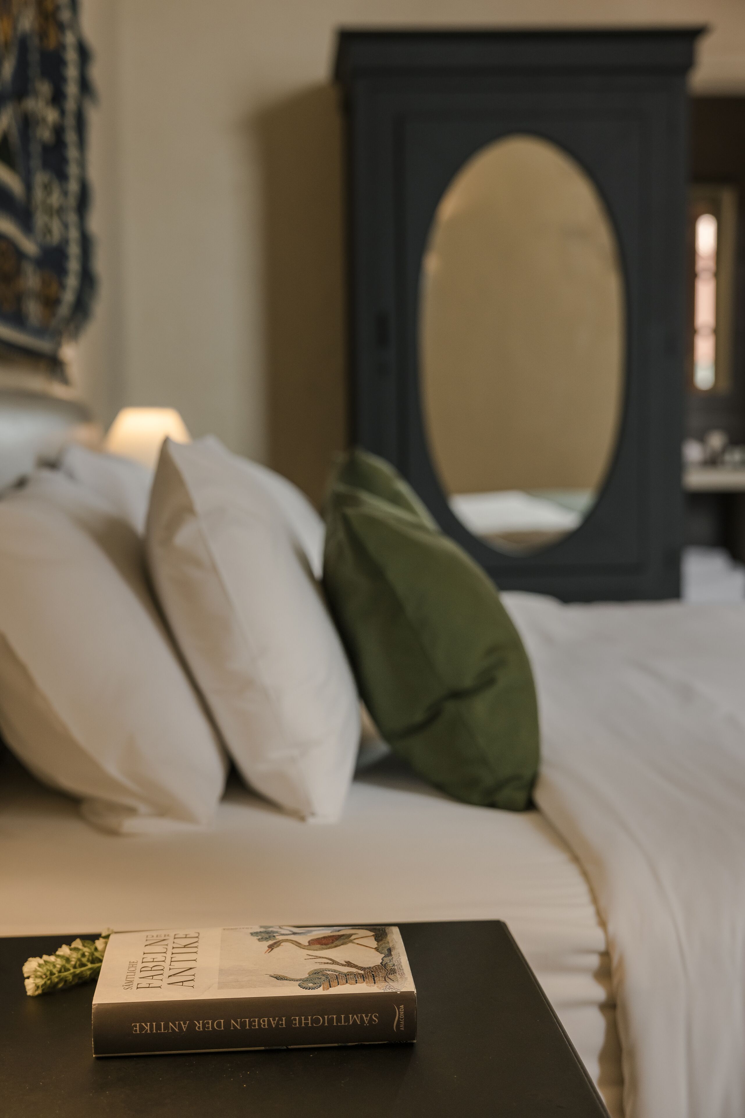 Cama bem arrumada com um livro intitulado "The Art of War" e um pequeno buquê de flores em uma mesa de cabeceira.