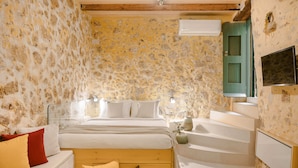 Quarto com parede de pedra, cama com roupa de cama branca, um banco de madeira e uma porta verde.