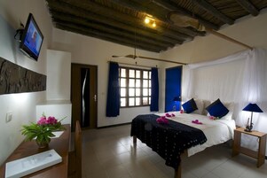 Quarto com cama, escrivaninha, cadeira, janela com cortinas, ventoinha no teto e televisão na parede.