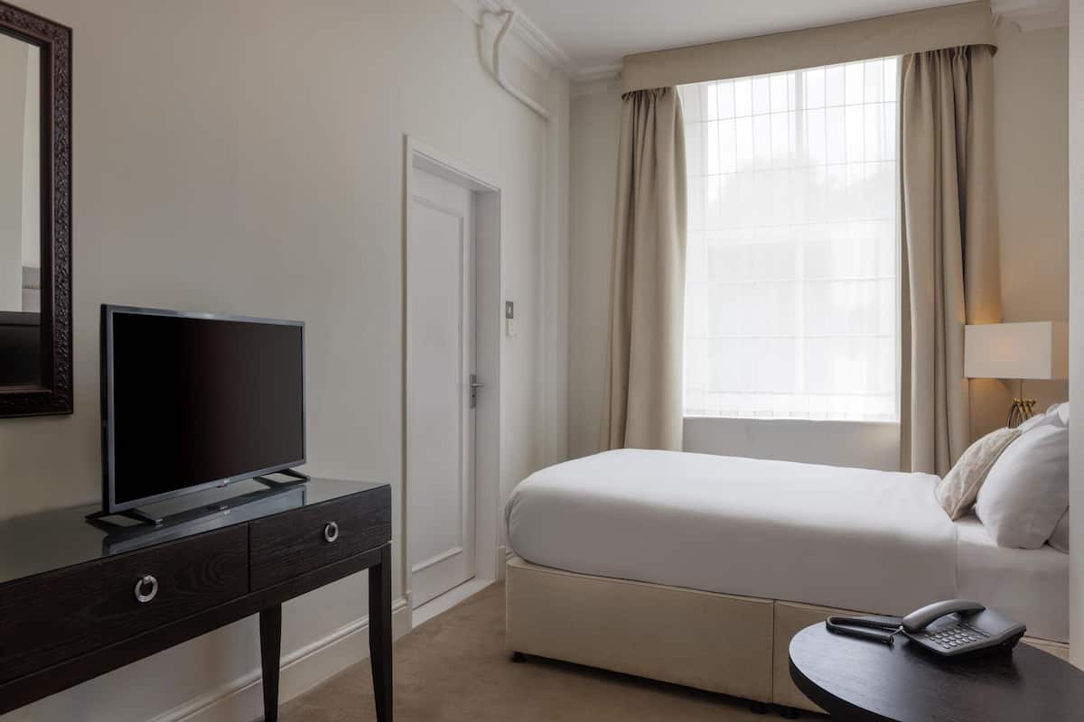 Quarto de hotel com uma cama, uma televisão sobre uma escrivaninha de madeira escura, um telefone num puzinho e uma janela com cortinas.