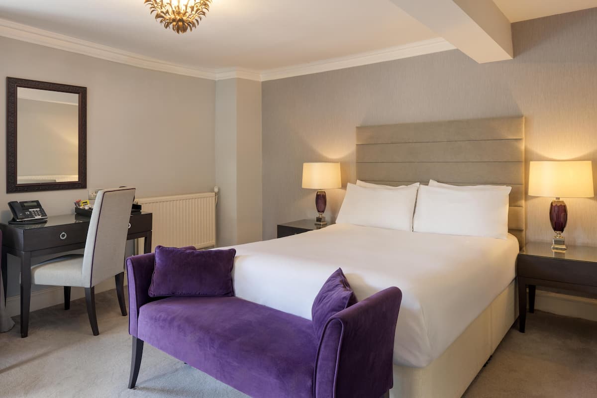 Quarto de hotel com uma cama grande, duas mesas de cabeceira com luminárias, uma escrivaninha com telefone e uma cadeira roxa.