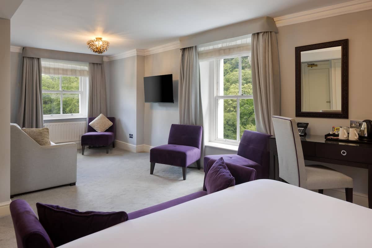 Quarto de hotel com uma cama grande, duas poltronas roxas, um sofá cinzento e uma escrivaninha com um espelho. Há também uma televisão fixada na parede.