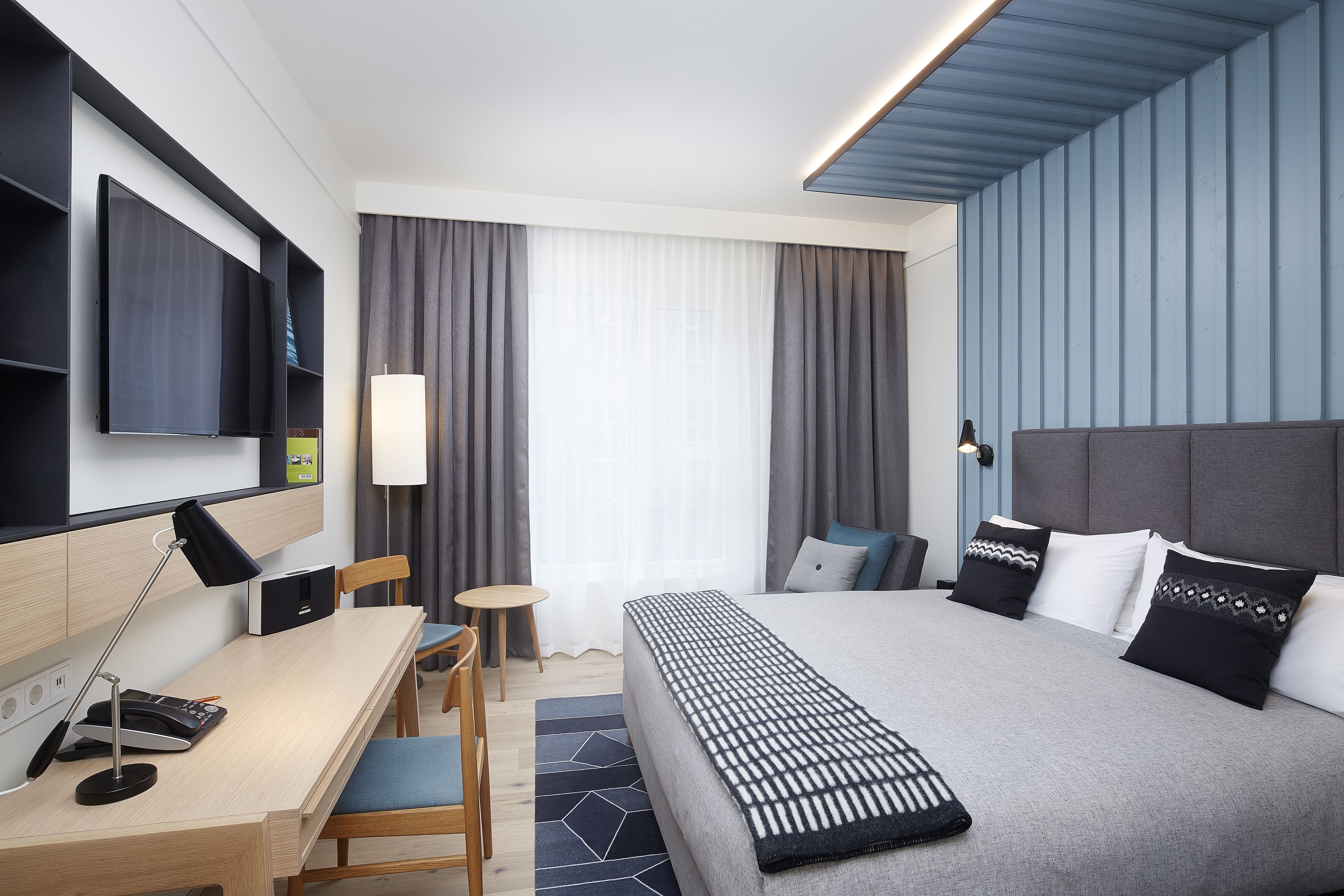 Quarto de hotel moderno com televisão de tela plana, uma escrivaninha com luminária, cama com colcha estampada e vista para o exterior por grandes janelas.
