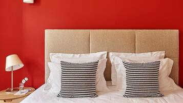 Quarto de hotel com parede vermelha, cabeceira bege, duas almofadas brancas e uma pequena mesa de cabeceira com uma luminária e um copo d'água.