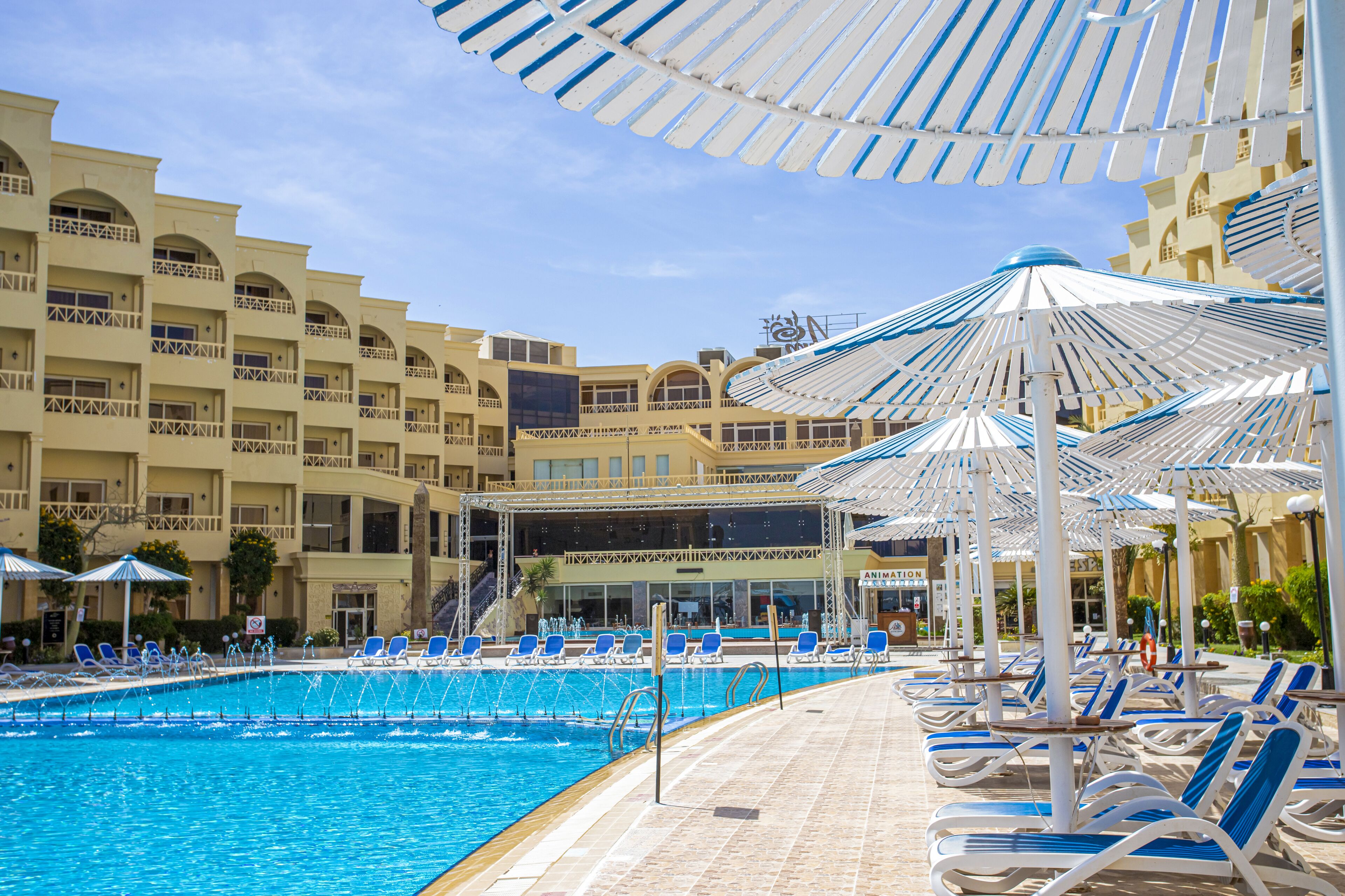 Piscina com espreguiçadeiras e guarda-sóis, rodeada por edifícios de hotel com vários andares.