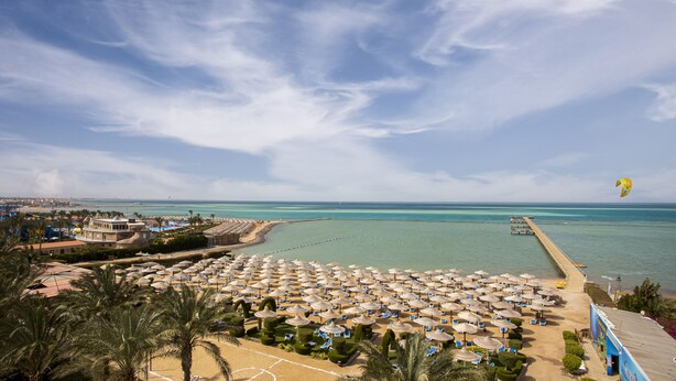 Um resort à beira-mar com fileiras de guarda-sóis e espreguiçadeiras, um cais que se estende para o mar, e um céu claro.