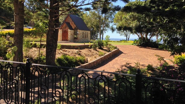 Um edifício de pedra com porta de madeira e telhado inclinado, rodeado por árvores e um caminho pavimentado.