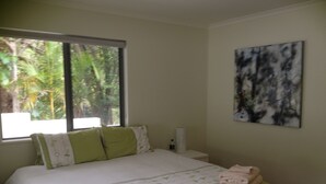 Quarto com cama, almofadas, mesa de cabeceira, abajur e janela com vista para o verde.