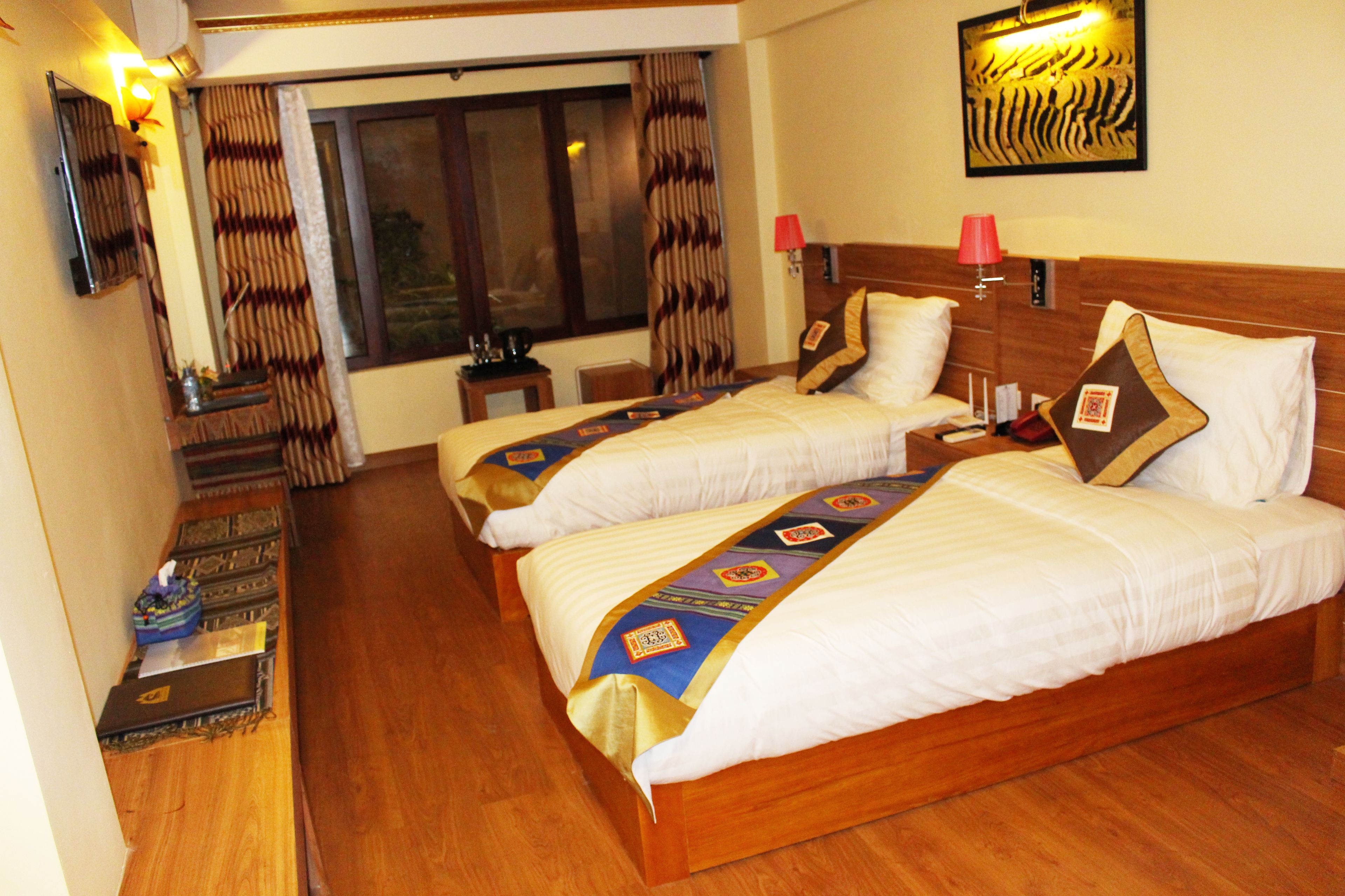 Quarto de hotel com duas camas, piso de madeira, uma janela grande com cortinas, uma obra de arte na parede e uma mesa de cabeceira com abajur.