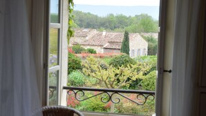 Quarto com cama, uma cadeira de vime e janela com vista para um jardim e edifícios.