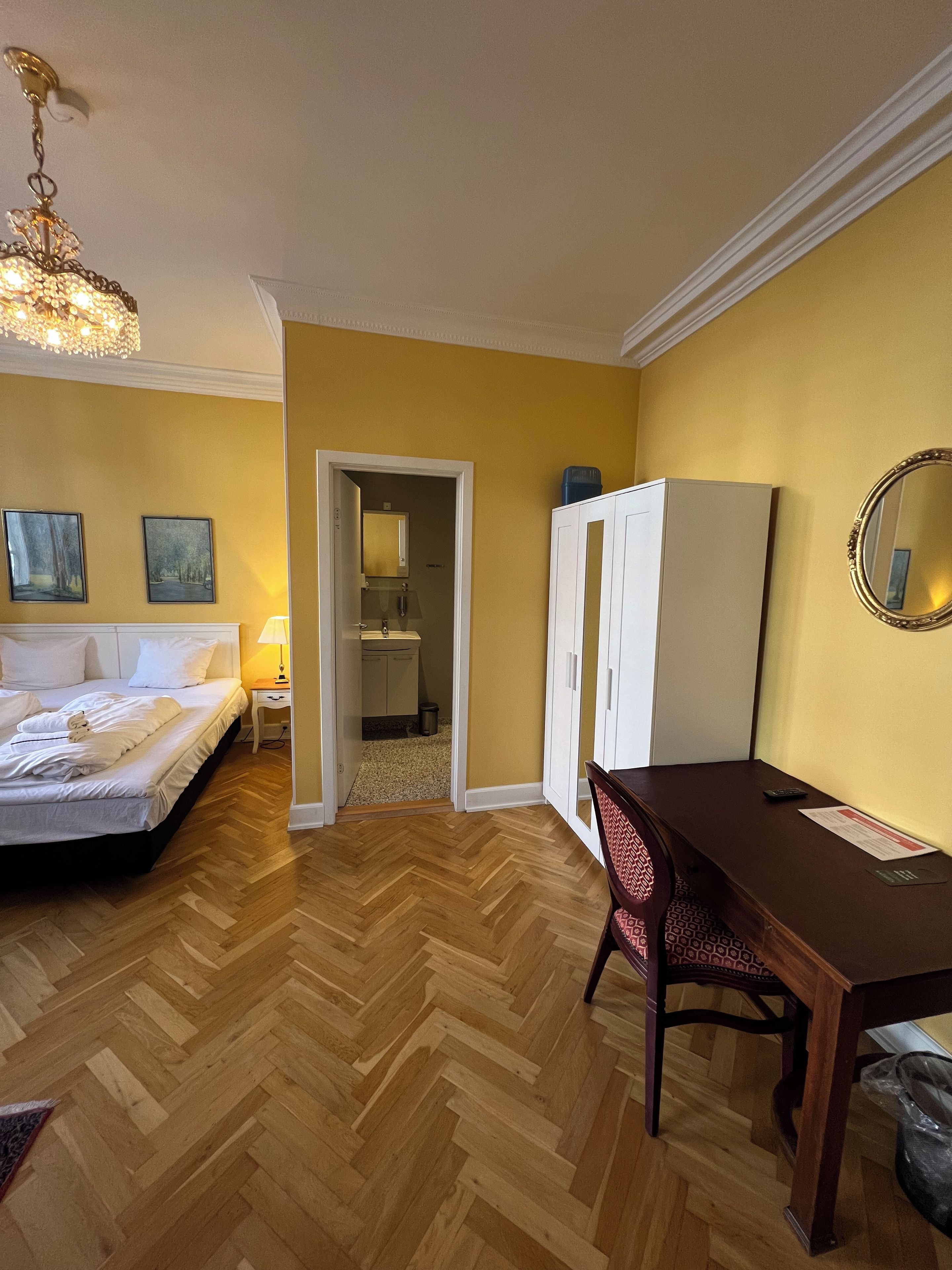 Quarto de hotel com cama, escrivaninha, cadeira, guarda-roupa, espelho e candelabro.