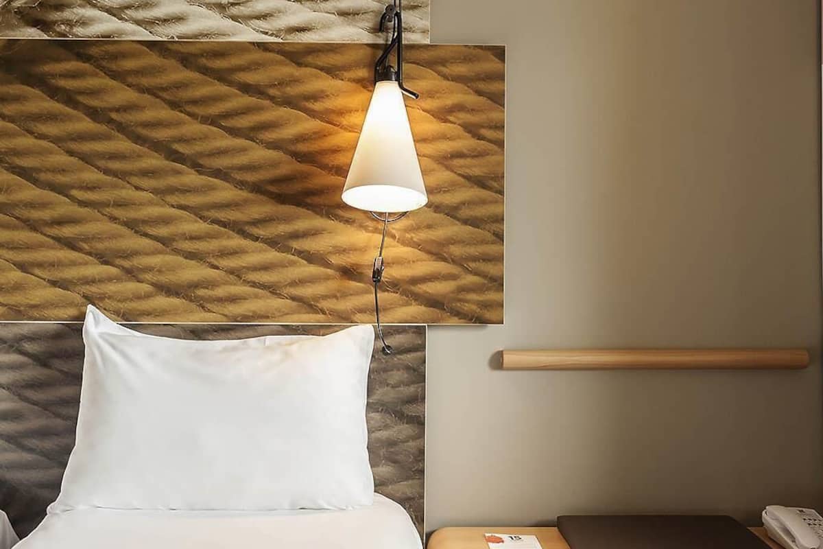 Quarto de hotel com cama, mesa de cabeceira com abajur, cabeceira de madeira e telefone fixado na parede.