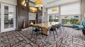 Uma ampla sala de jantar com uma grande mesa de madeira, cadeiras pretas e um tapete estampado. Há duas luminárias pendentes, uma lareira e uma janela grande com persianas.